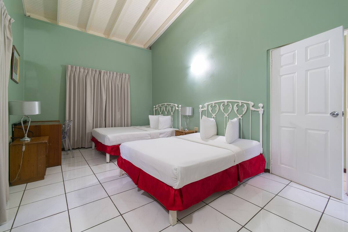 Quarto com duas camas, cômoda de madeira, espelho, porta e janela com cortinas.
