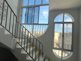 Escadaria com corrimão de metal que leva a um patamar com grandes janelas, oferecendo vista de edifícios e de uma grua.
