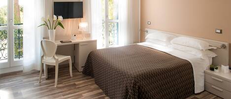 Quarto de hotel com cama, escrivaninha com cadeira, televisão e janela com cortinas.