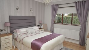 Quarto com cama grande, luminárias de cabeceira, cômoda, janela com cortinas e lustre.