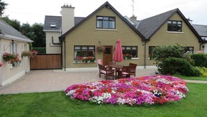 Uma casa de dois andares com fachada de tijolos, um jardim da frente com flores coloridas e um alpendre com móveis de exterior.