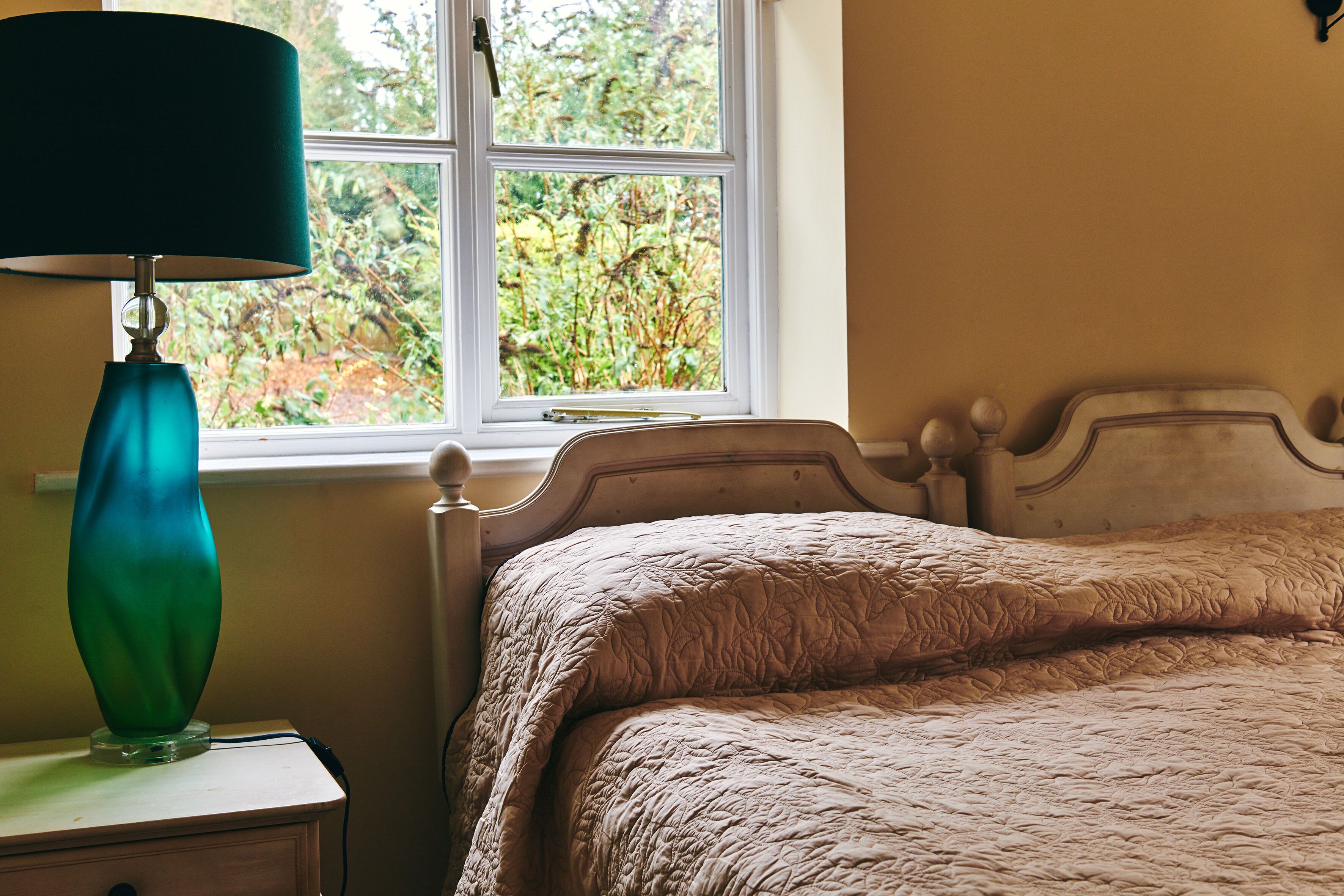 Quarto com cama, mesa de cabeceira com luminária e janela com vista para o verde.