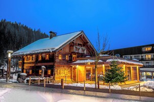Um chalé de madeira com telhado coberto de neve e interior iluminado, rodeado por neve e árvores.