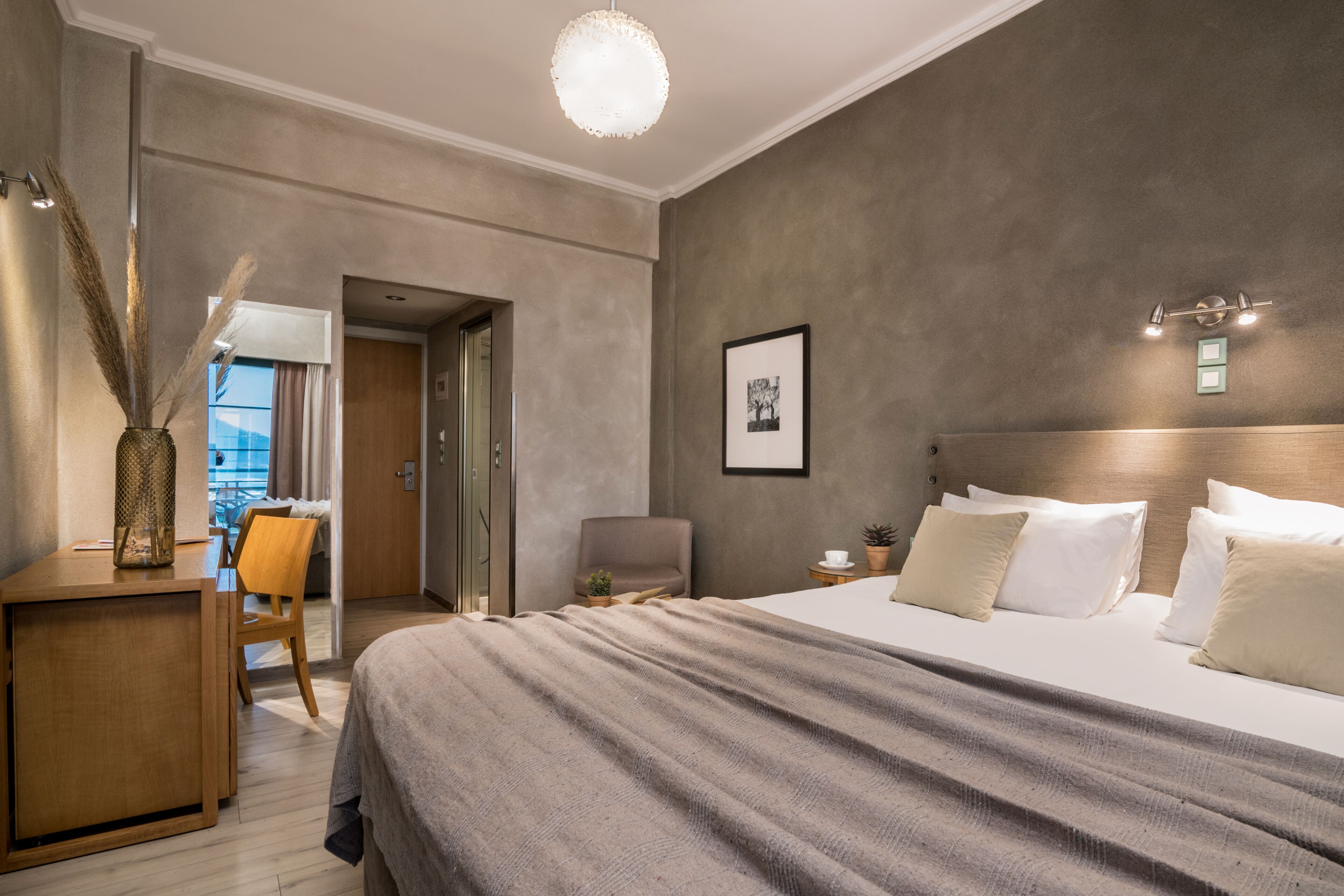 Quarto de hotel moderno com uma cama grande, uma escrivaninha e uma cadeira. Há uma porta que dá para outro quarto e uma janela com cortinas.