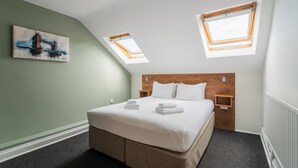 Quarto com uma cama grande, duas mesas de cabeceira, cabeceira de madeira e duas claraboias.