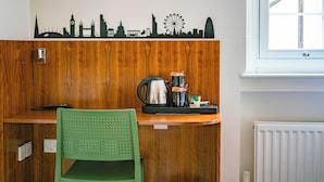 Divisória de madeira com silhueta de um horizonte urbano, uma cadeira verde, um espaço para café com chaleira, caneca e petiscos, e uma janela com persianas.