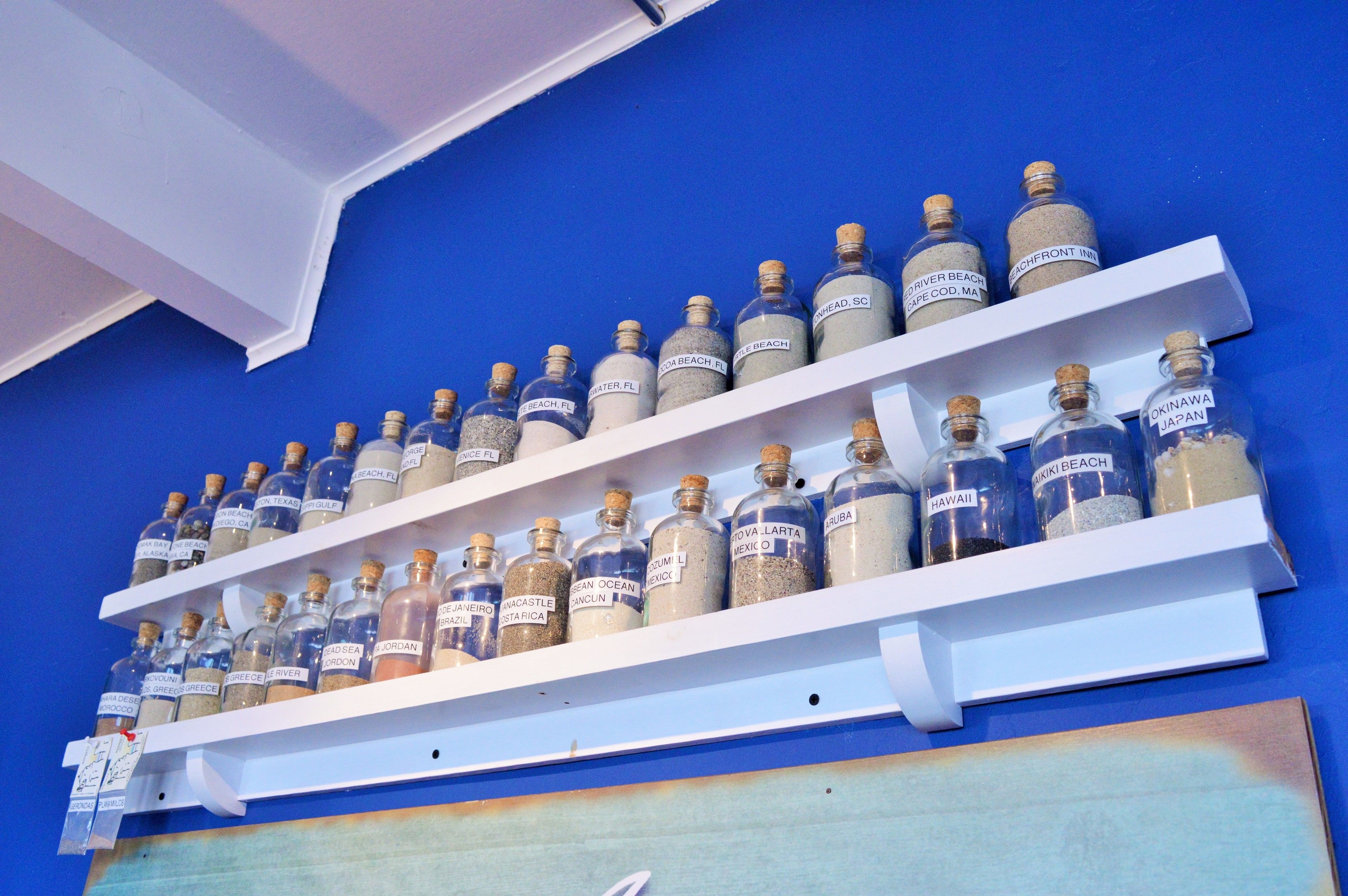 Exposição de garrafas com etiquetas mostrando diferentes nomes de praias, em prateleiras brancas contra uma parede azul.