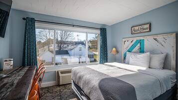Quarto com uma janela grande, uma cama com cabeceira branca e azul, e vista para uma casa lá fora.