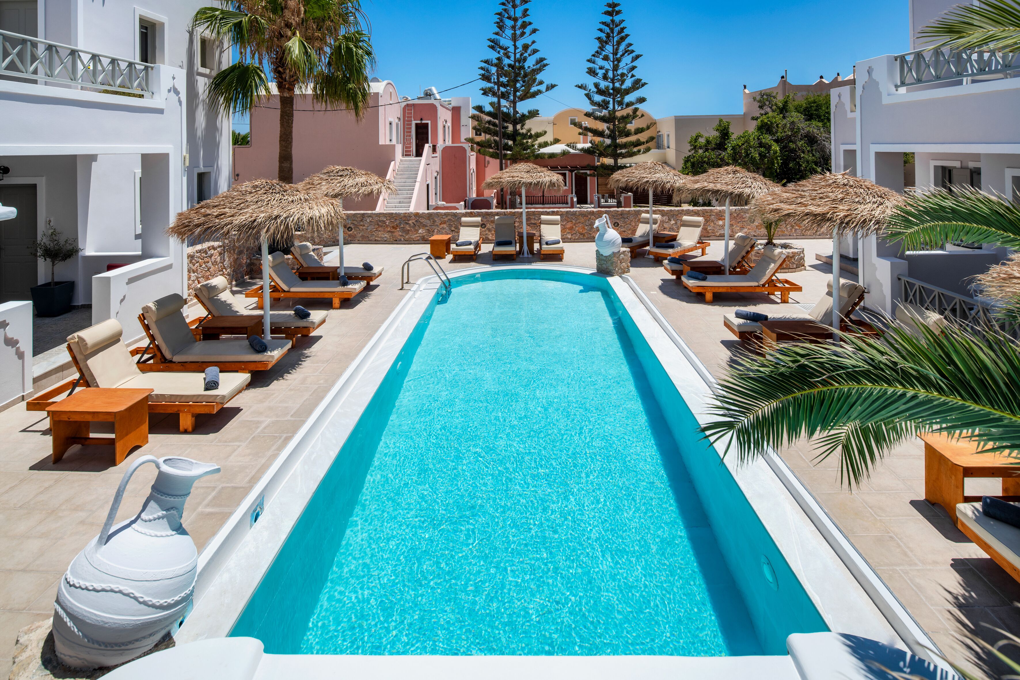 Piscina com espreguiçadeiras e guarda-sóis, cercada por edifícios com fachadas brancas e cor-de-rosa.