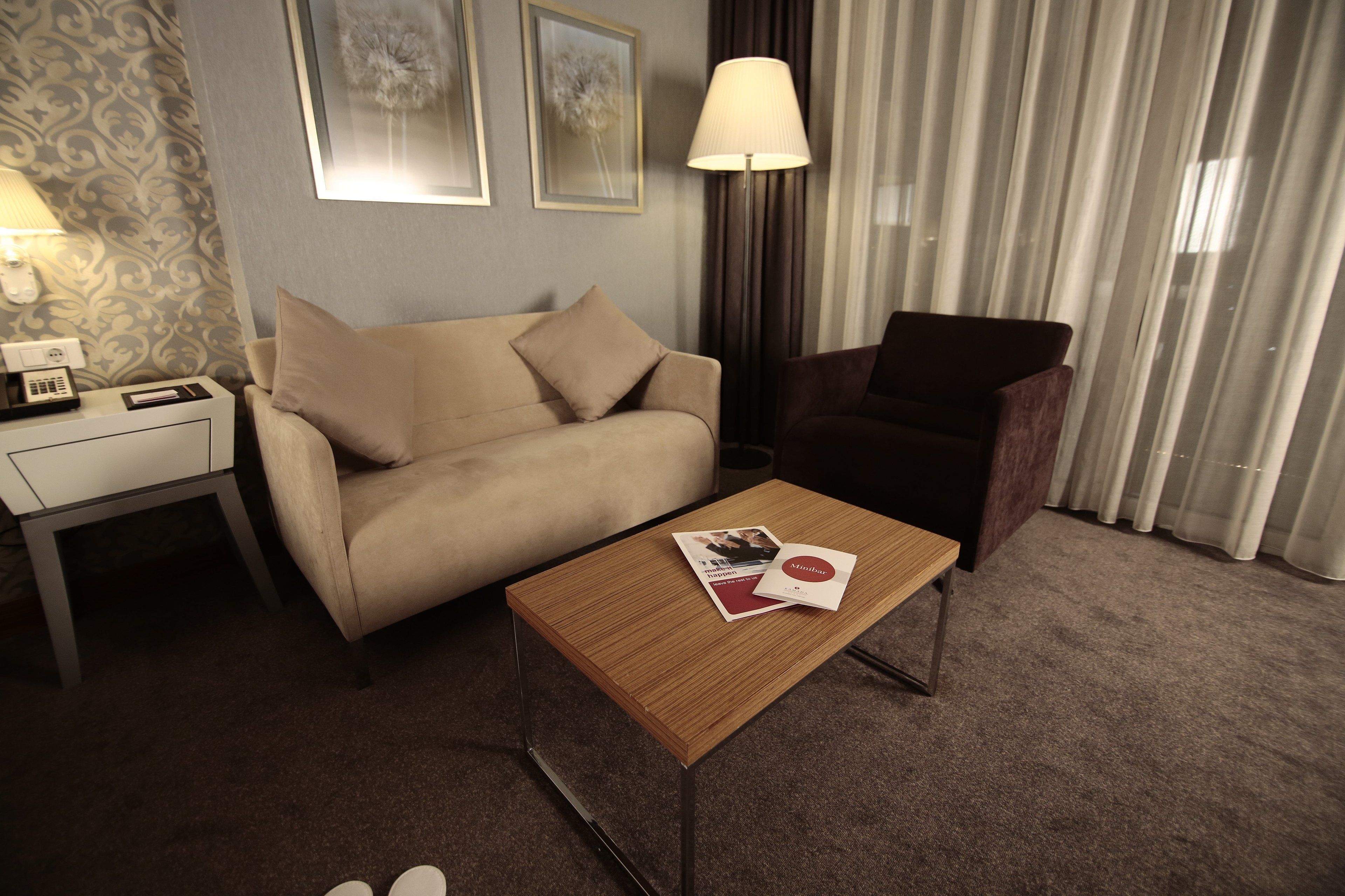 Quarto de hotel com sofá bege, poltrona escura, mesa de centro de madeira com revistas, luminária e mesa de cabeceira com telefone e luminária.