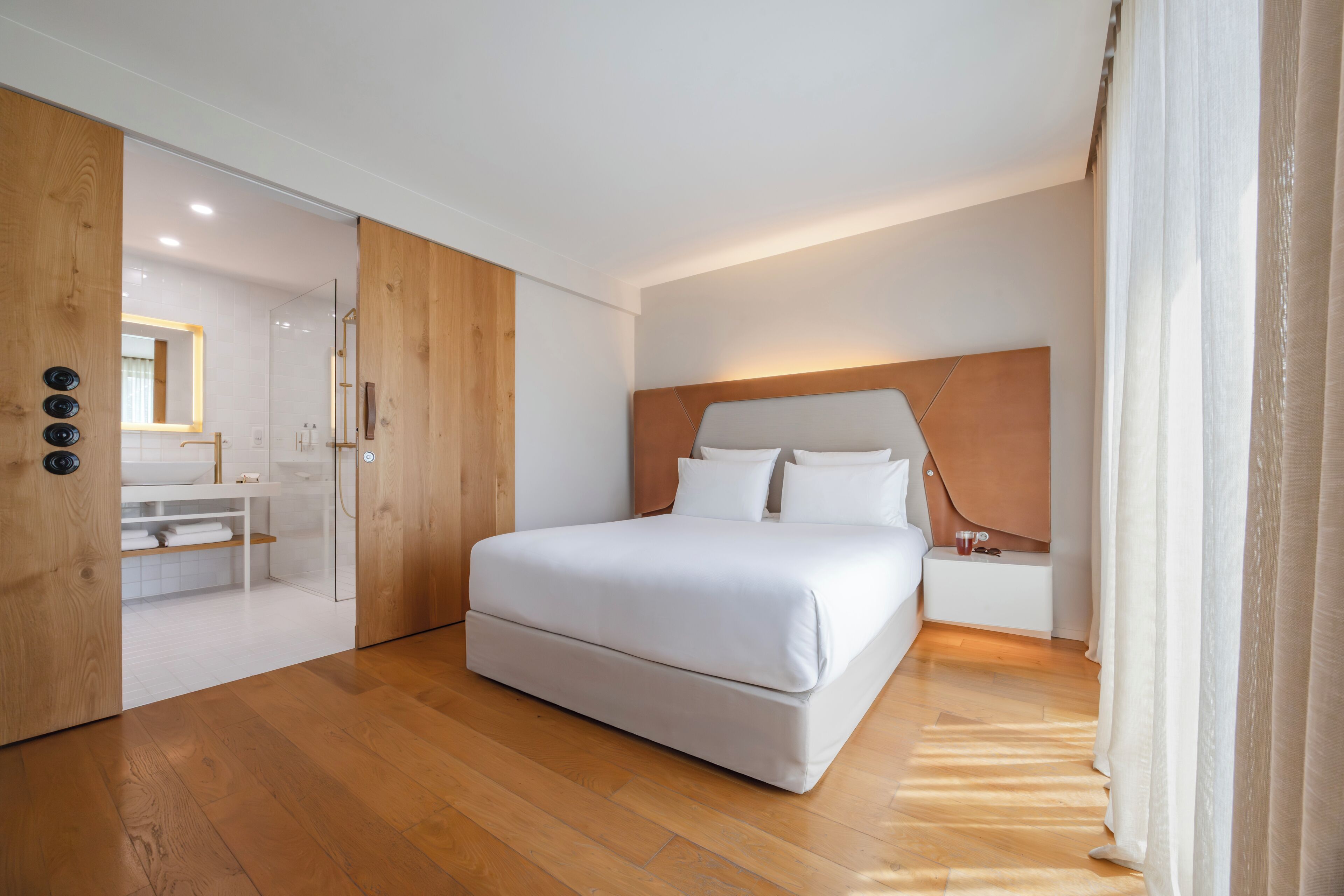 Quarto de hotel moderno com uma cama grande, cabeceira de madeira, mesa de cabeceira com abajur e banheiro visível através de uma porta deslizante.