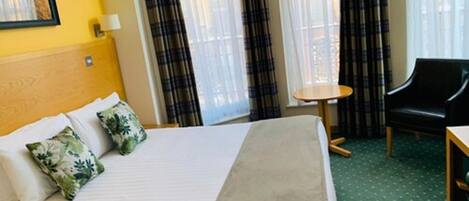 Quarto de hotel com cama, poltrona, mesinha e cortinas.