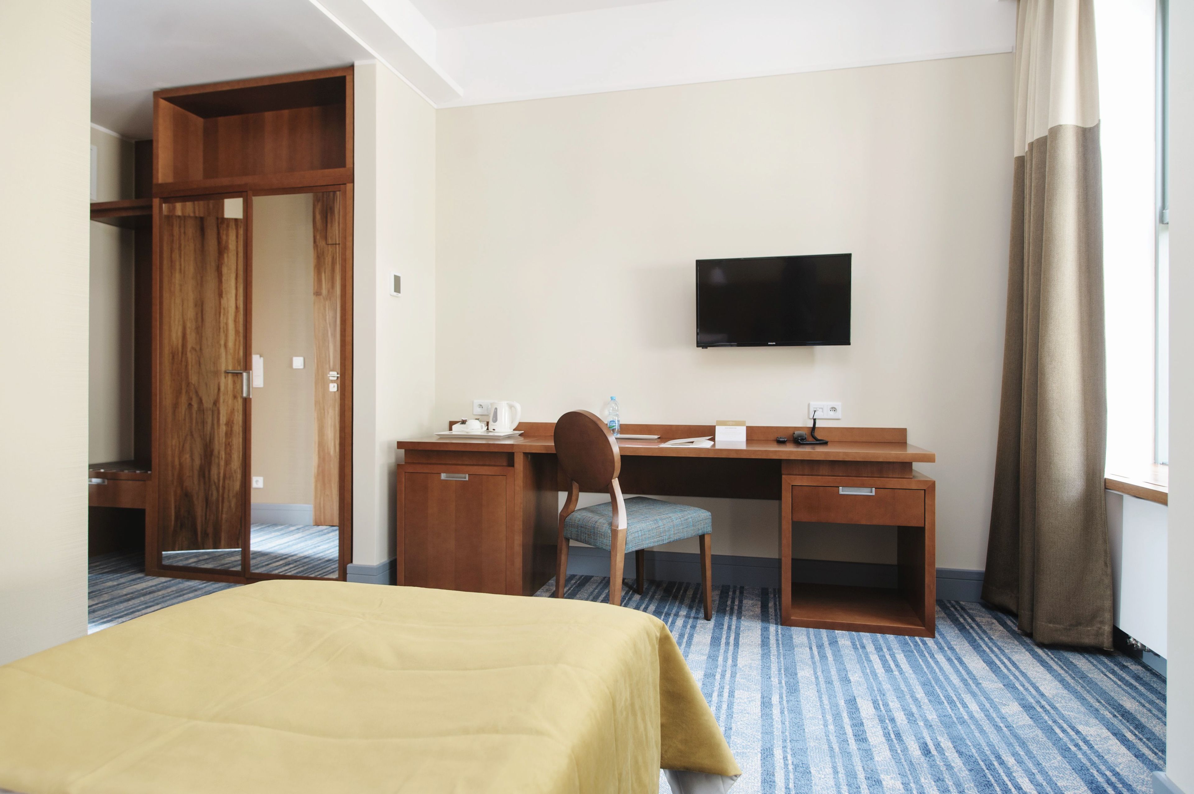Quarto de hotel com cama, escrivaninha, cadeira, televisão e guarda-roupa.