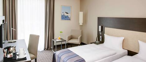 Quarto de hotel com duas camas, uma escrivaninha com cadeira, uma mesinha de cabeceira com abajur e um quadro pendurado na parede.