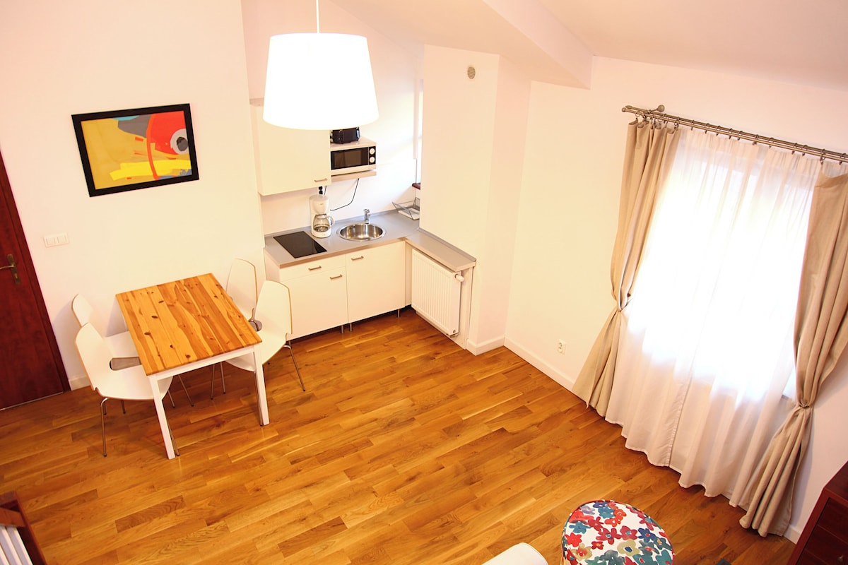 Cozinha compacta com mesa de madeira, cadeiras brancas, luminária pendente e janela com cortinas.