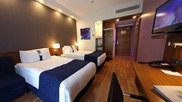 Quarto de hotel com duas camas, uma escrivaninha, televisão e piso de madeira.