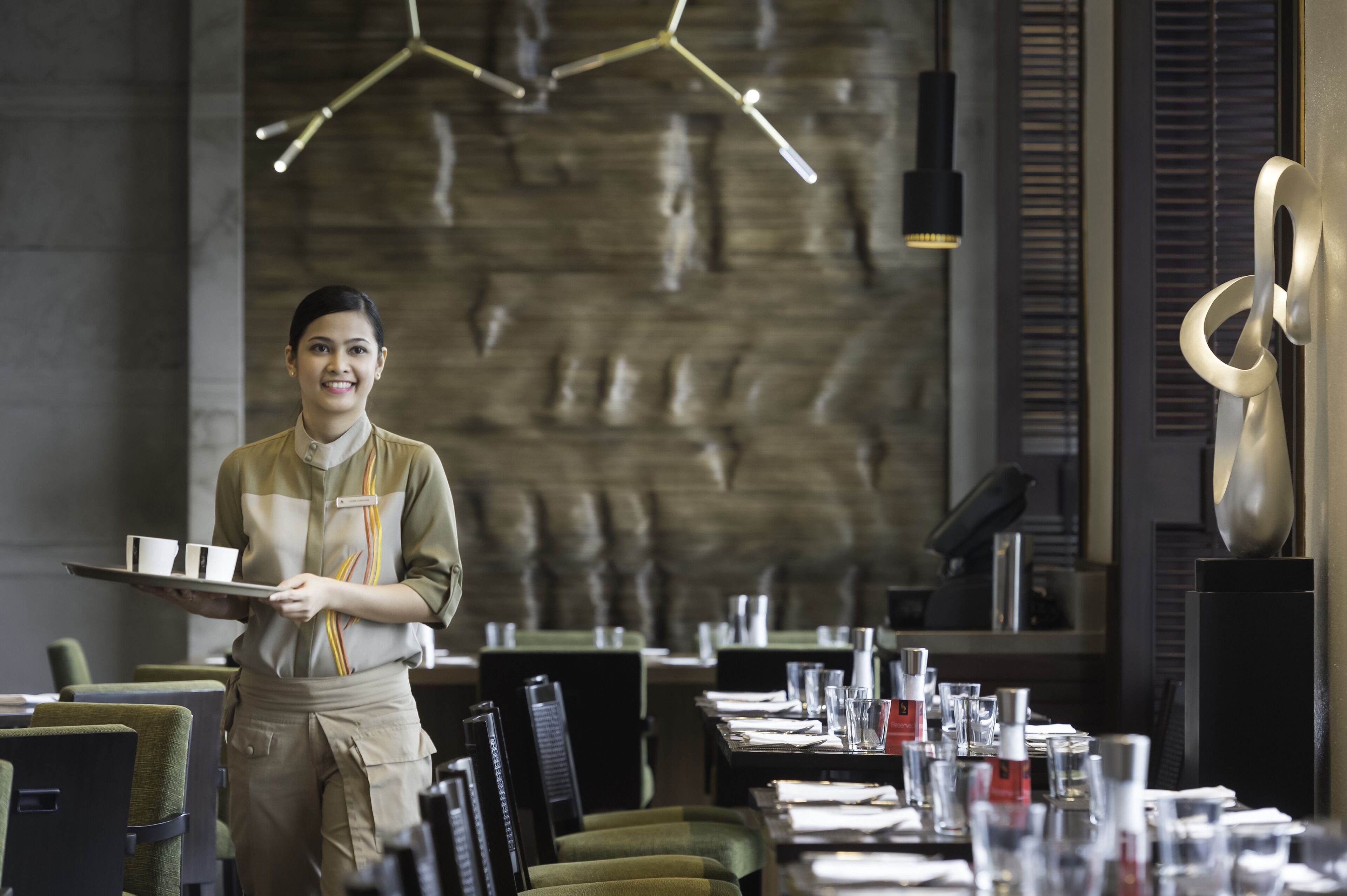 Interior de restaurante com um garçom carregando uma bandeja, mesas montadas com copos e talheres, e um lustre moderno.