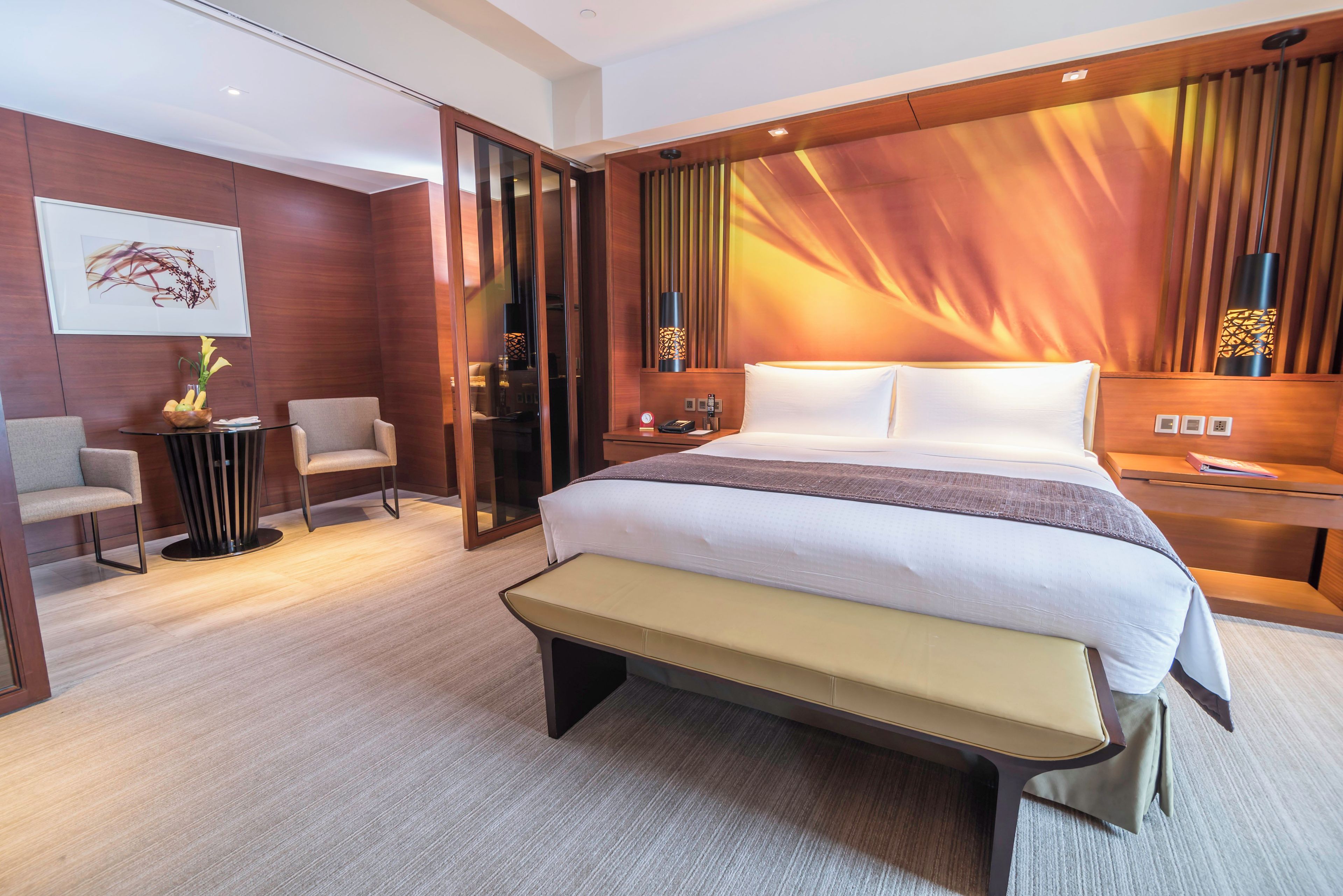 Quarto de hotel com uma cama grande, uma mesa de cabeceira, uma cadeira, uma pequena mesa com um vaso e uma parede com um quadro.