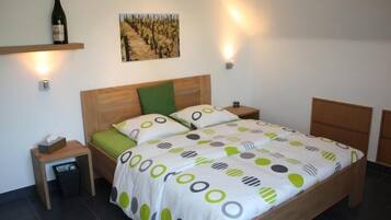 Quarto com cama de madeira, roupa de cama verde e branca, mesa de cabeceira com abajur, cesto de lixo e prateleira na parede com uma garrafa.