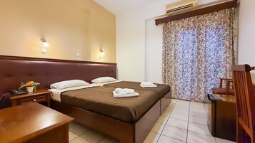 Quarto de hotel com uma cama grande, uma escrivaninha e uma cadeira. Há uma janela com cortinas e um ar condicionado acima.