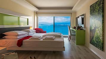 Quarto de hotel moderno com uma cama, uma escrivaninha, uma cadeira, uma televisão e uma grande janela com vista para o mar.