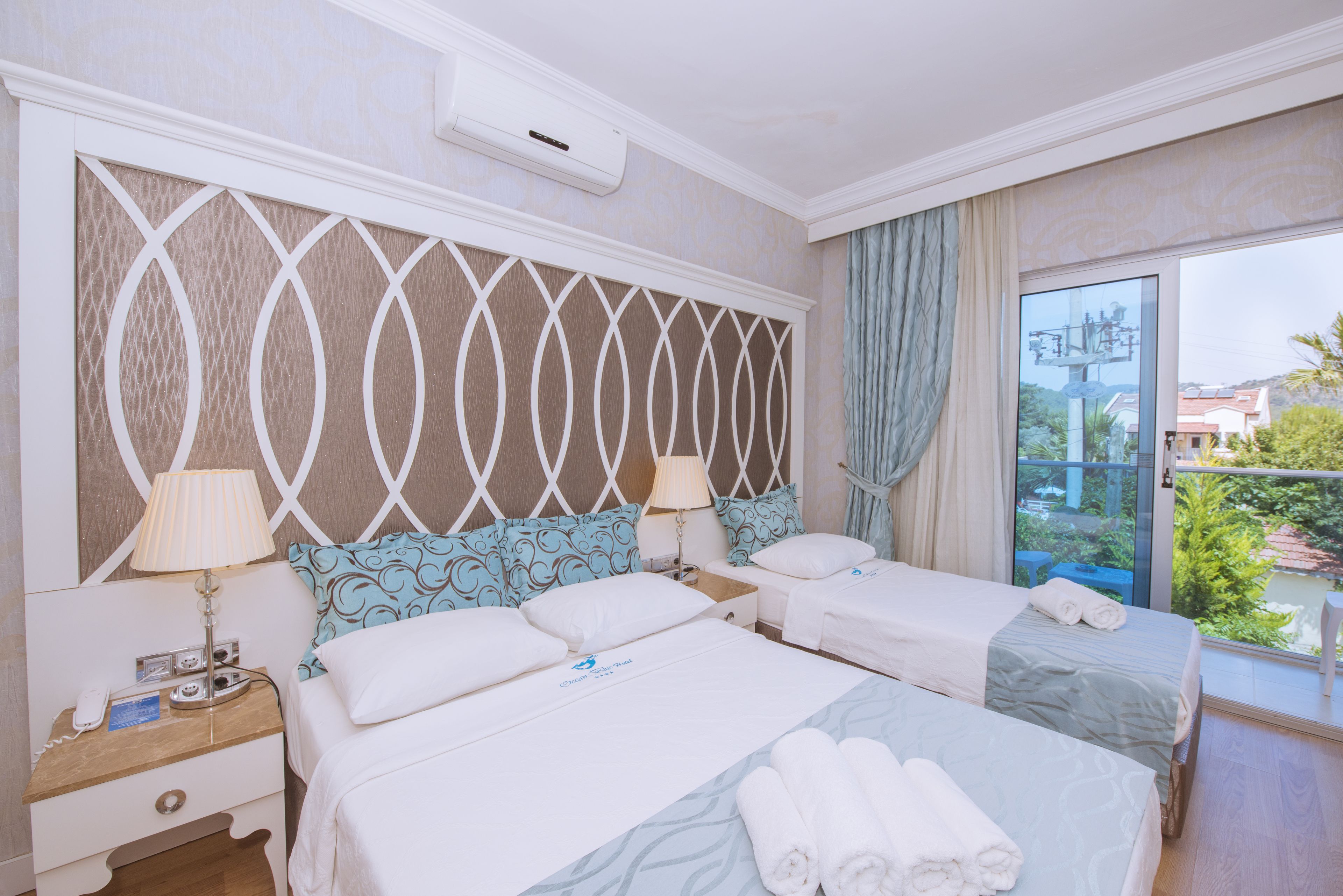 Quarto de hotel com duas camas, uma janela grande com vista, cabeceira decorativa e mesa de cabeceira com luminária e telefone.