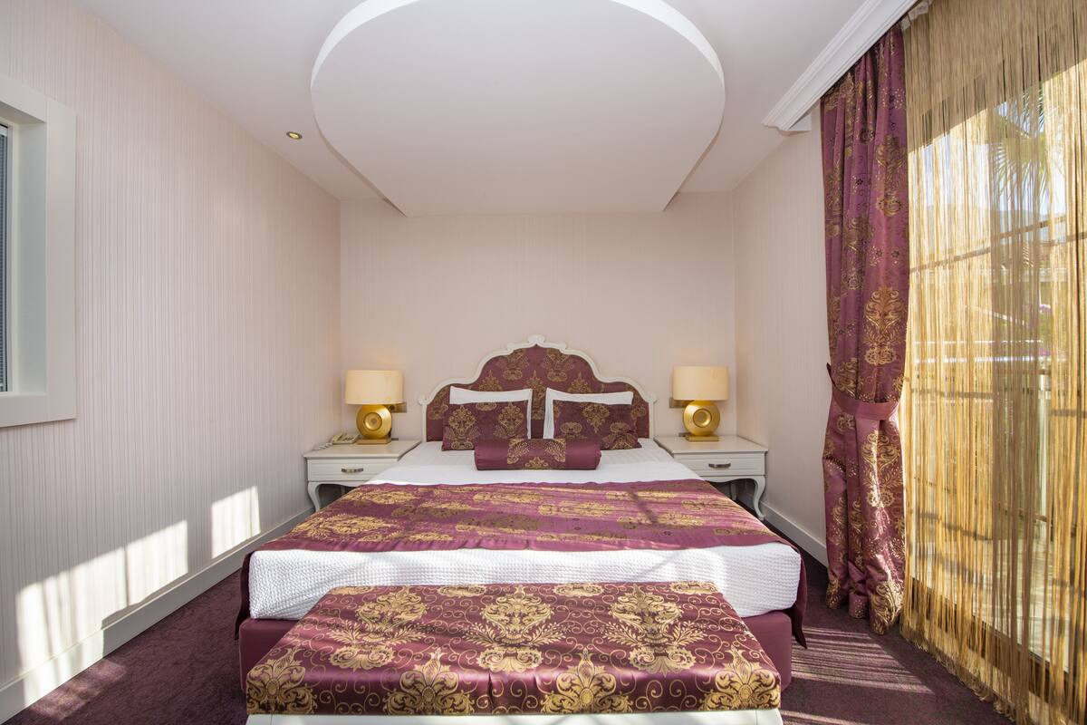 Quarto de hotel com uma cama grande, duas mesas de cabeceira com abajures, uma janela com cortinas e uma luminária no teto.