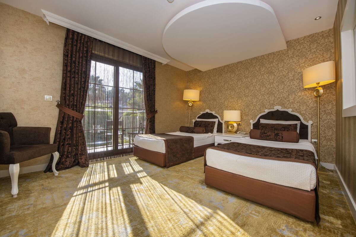 Quarto de hotel com duas camas, um sofá, uma escrivaninha e uma janela grande com cortinas.