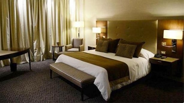 Quarto de hotel com uma cama grande, duas mesas de cabeceira com luminárias, uma cadeira, uma mesinha e uma janela ampla com cortinas.