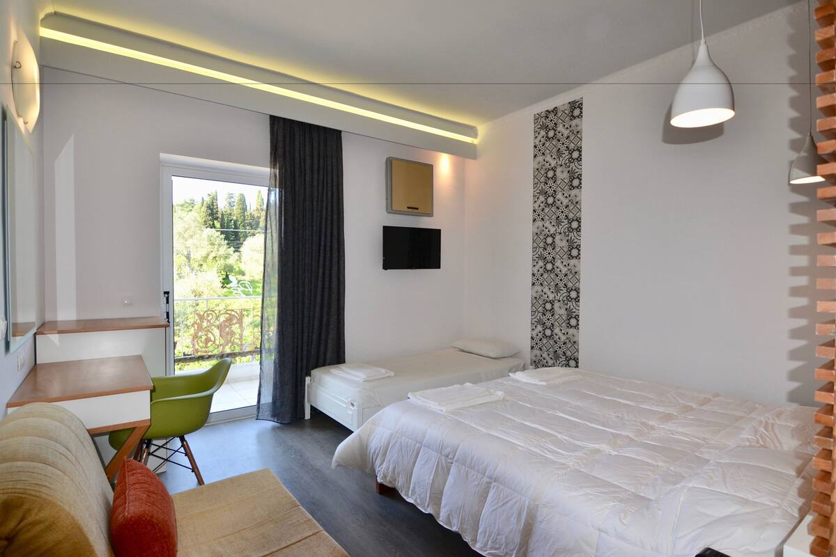 Quarto de hotel moderno com cama grande, escrivaninha, cadeira e varanda com vista para árvores.