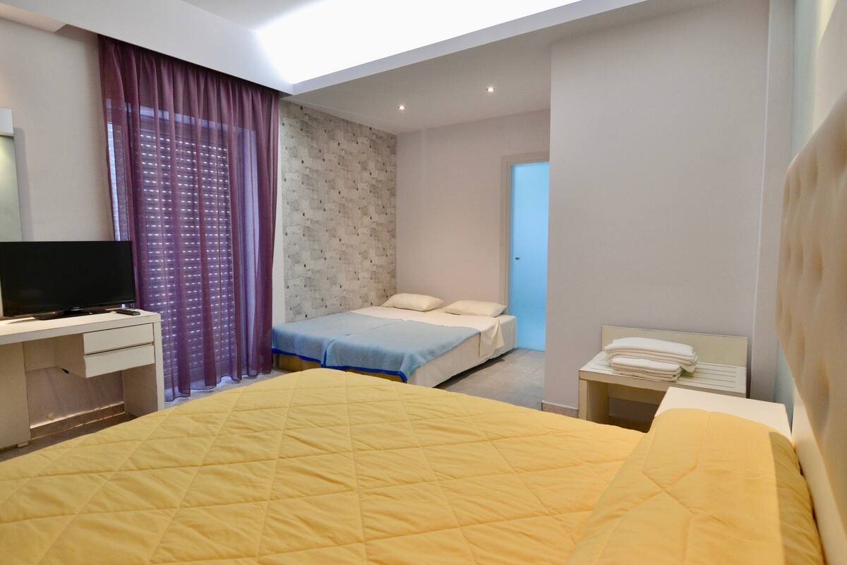 Quarto de hotel com uma cama grande, uma escrivaninha com televisão de tela plana, uma mesinha de cabeceira com toalhas e uma janela com cortinas.