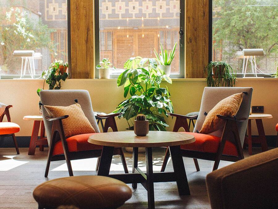 Um espaço de estar acolhedor com duas cadeiras e uma mesa, cercado por plantas em vasos e uma grande janela com vista para o verde.