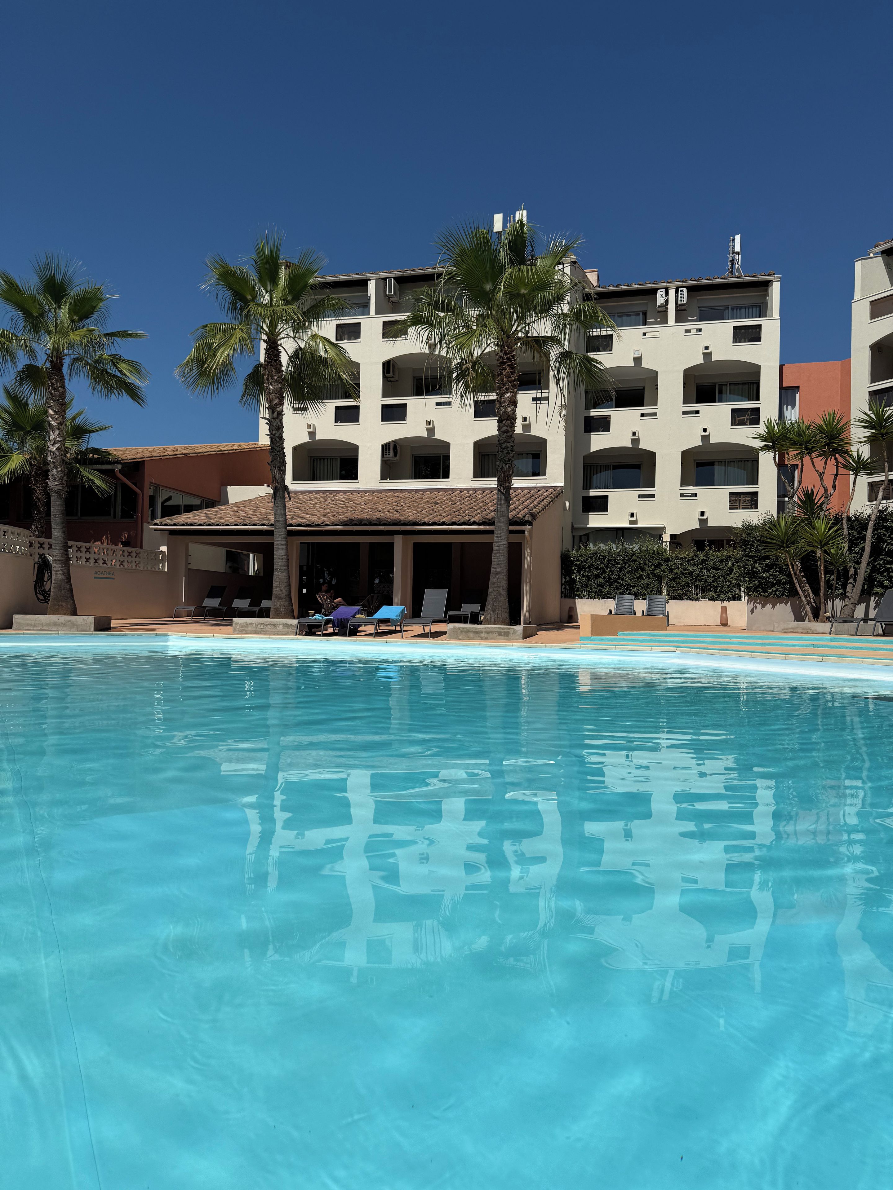 Piscina com superfície azul translúcida, rodeada por palmeiras e um edifício com varandas.