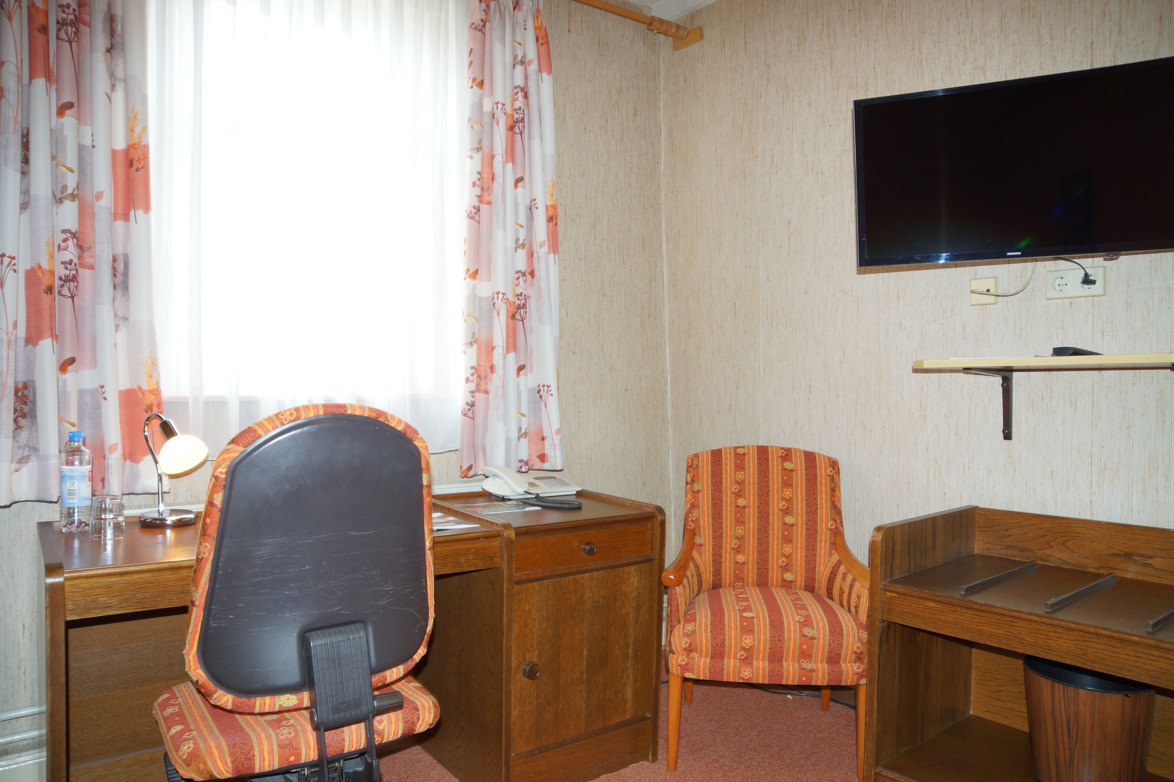 Quarto de hotel com secretária de madeira, uma cadeira, televisão na parede e janela com cortinas florais.