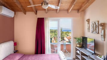 Quarto com cama, ventilador de teto, TV, porta de vidro deslizante para uma varanda e vista da paisagem.