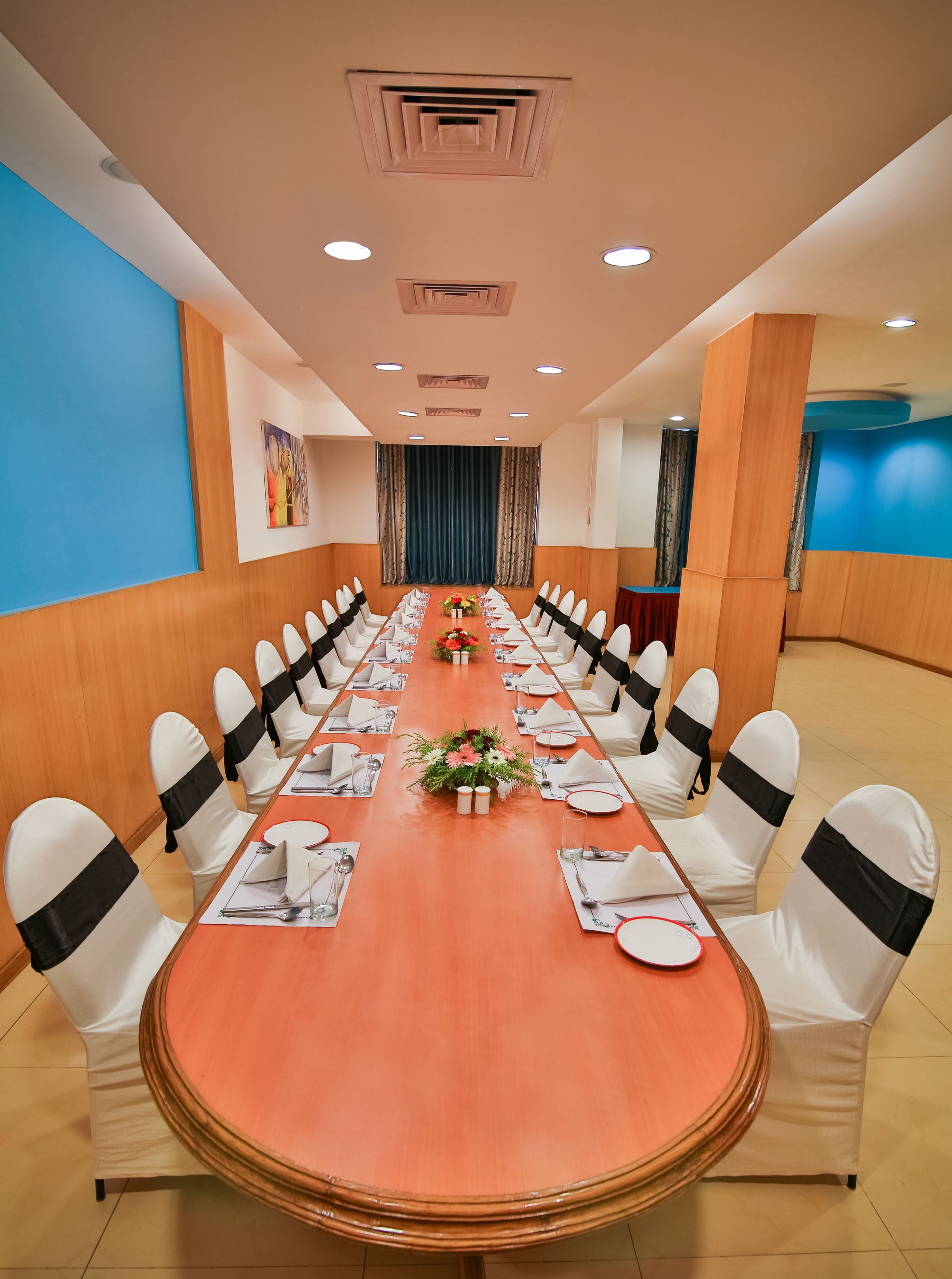 Sala de conferência com uma mesa comprida, cadeiras brancas com fitas pretas e uma parede de destaque azul.