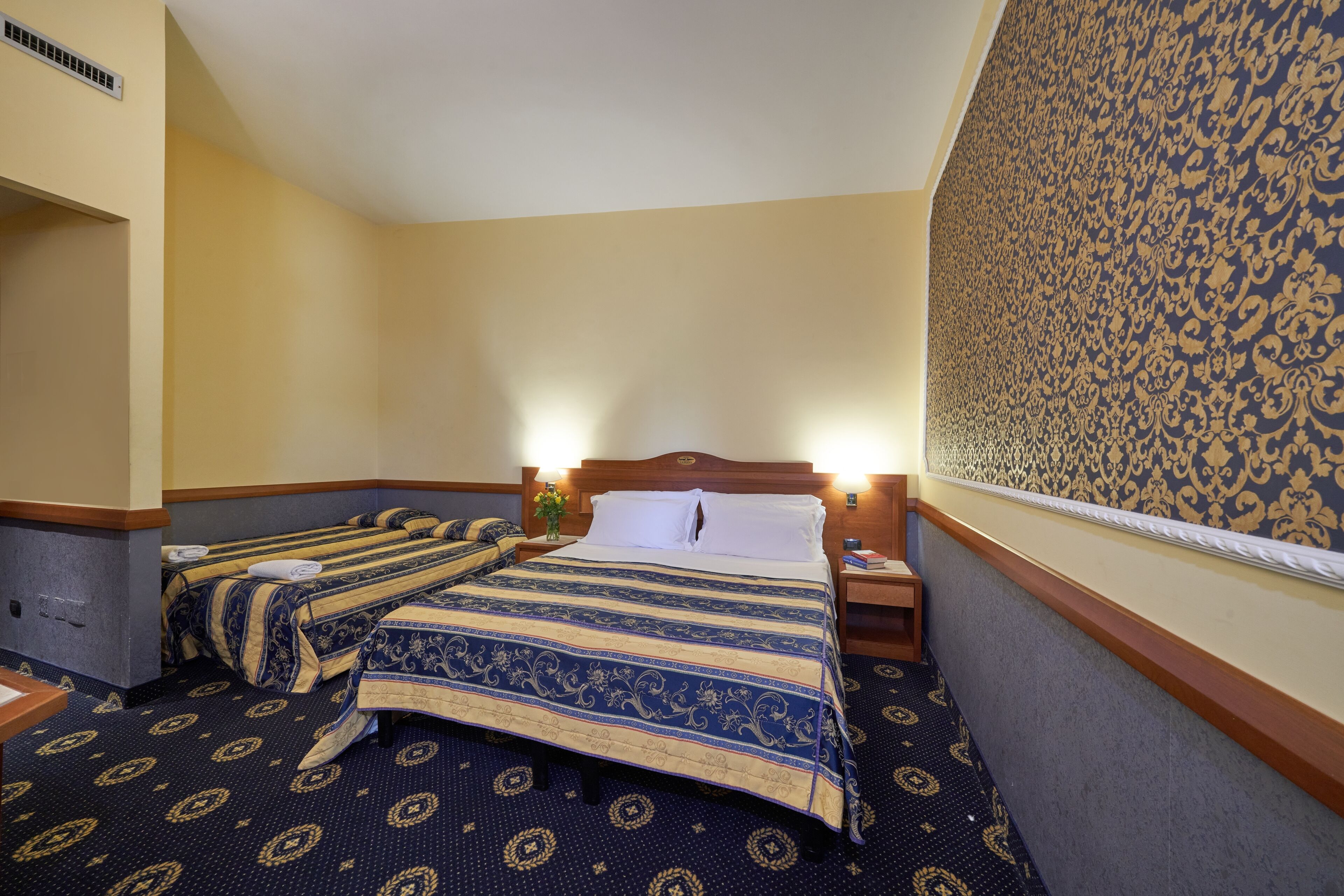Quarto de hotel com duas camas, cabeceira de madeira, mesa de cabeceira e abajur, e parede estampada.