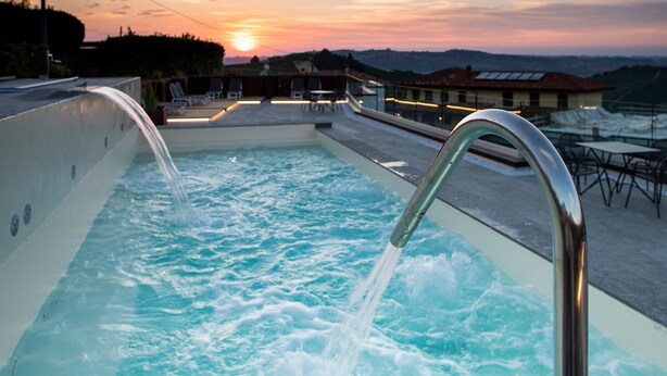Piscina com jatos de água, rodeada por espreguiçadeiras e mesas, com vista para o pôr do sol ao fundo.