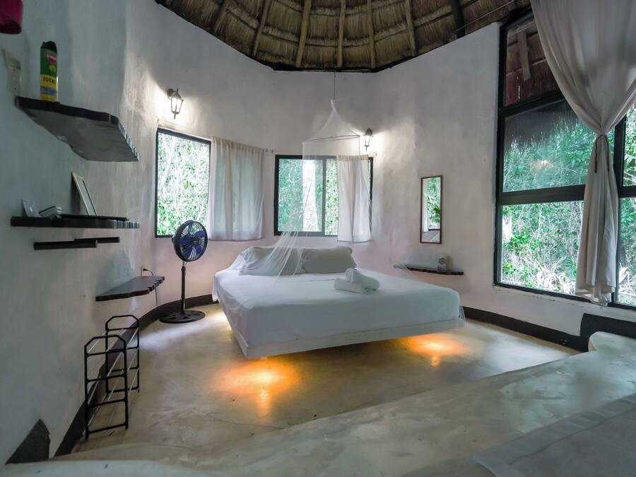 Quarto com cama, mesas de cabeceira, um ventilador e grandes janelas com cortinas.