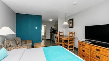 Quarto de hotel com cama, escrivaninha, cadeira, televisão e cozinha compacta ao fundo.
