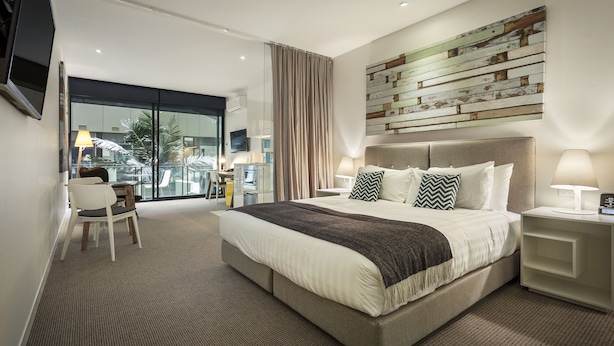 Quarto de hotel moderno com uma cama grande, televisão de tela plana, escrivaninha com cadeira e vista para palmeiras pela porta de vidro.