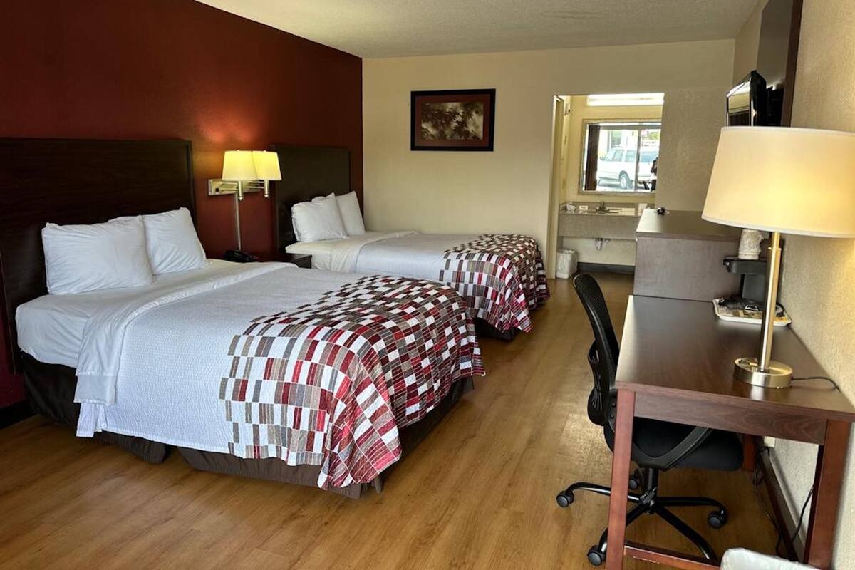 Quarto de hotel com duas camas, uma escrivaninha, uma cadeira, um abajur, um quadro e uma janela.