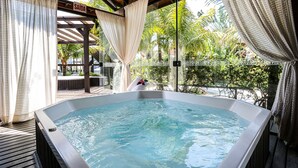 Uma banheira de hidromassagem com deck de madeira e estrutura de vidro, cercada por cortinas e plantas tropicais.