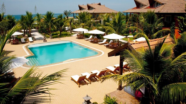 Área de piscina do resort com espreguiçadeiras e guarda-sóis, cercada por palmeiras e vista para o oceano.
