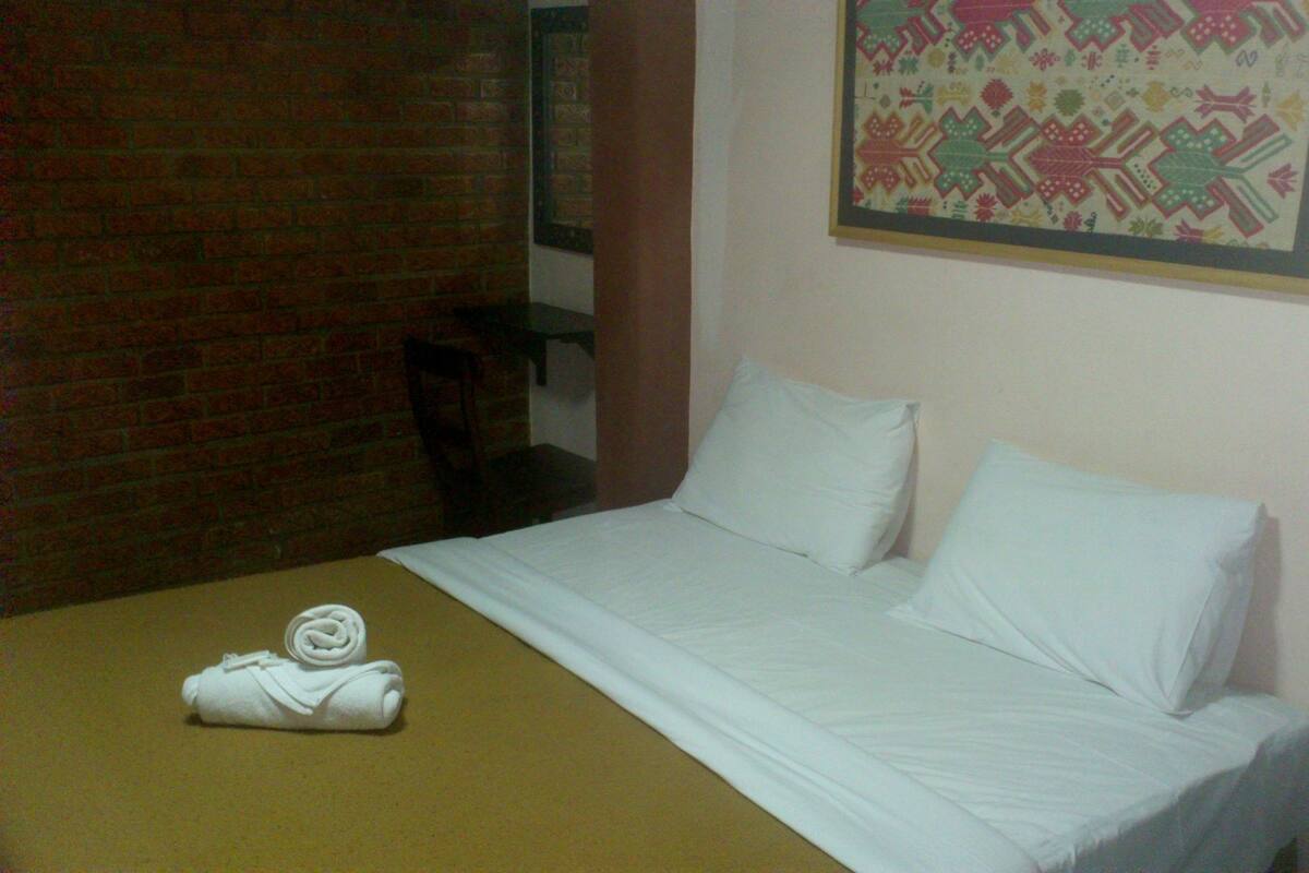 Cama com lençóis brancos e uma toalha enrolada, parede de tijolos e um pendurar colorido na parede.