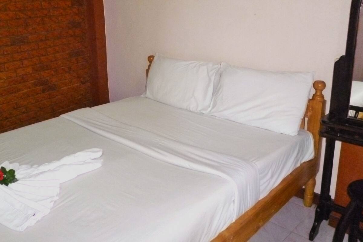 Uma cama de solteiro com roupa de cama branca, cabeceira de madeira e parede de tijolos ao fundo.