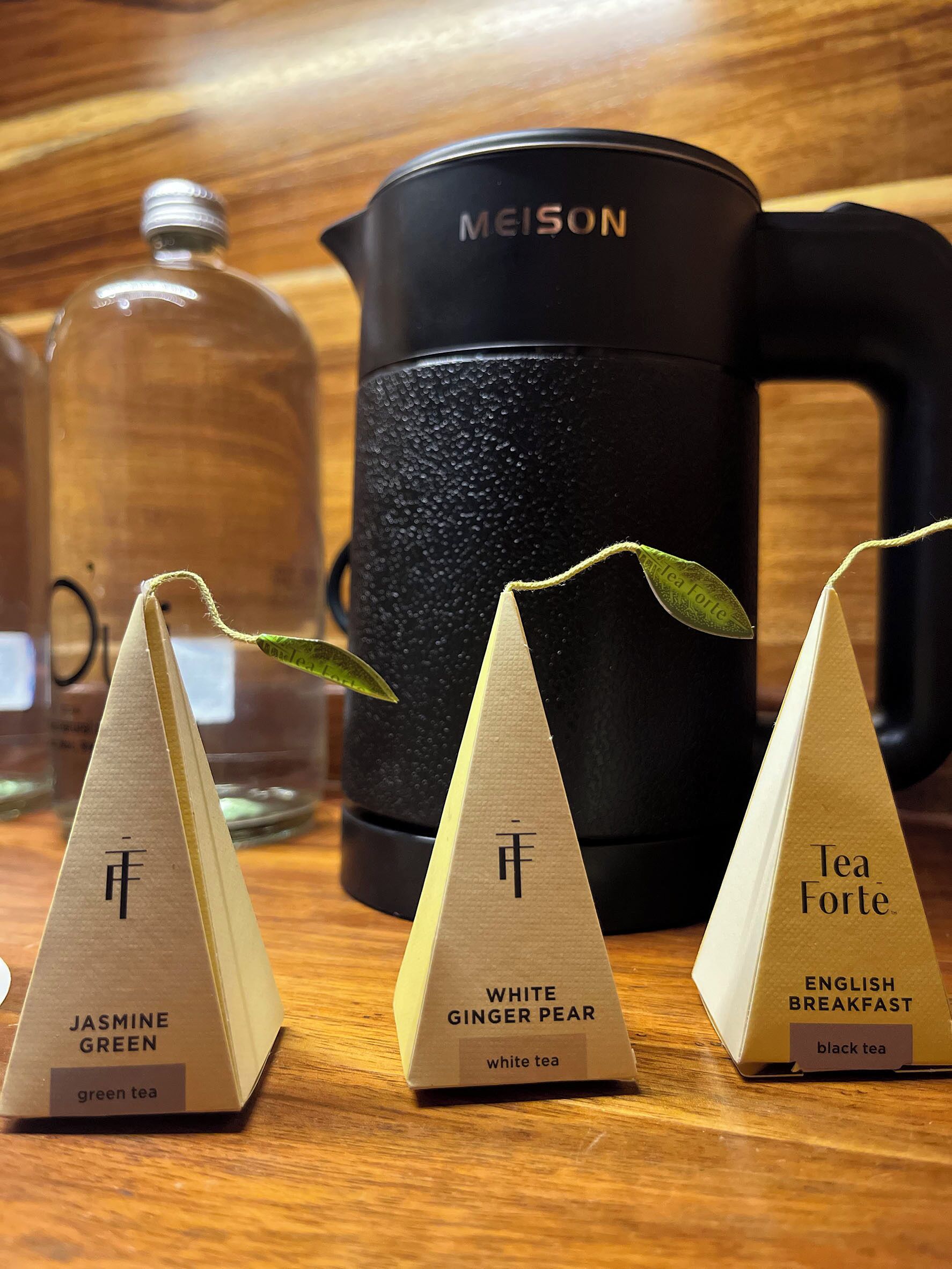 Três caixas de chá com as etiquetas 'Jasmine Green', 'White Ginger Pear' e 'Tea Forte' sobre uma superfície de madeira, com uma chaleira preta 'MEISON' ao fundo.