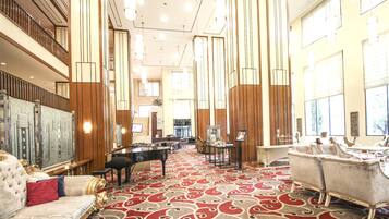 Um amplo hall de hotel com carpete estampado em vermelho e bege, um piano de cauda, um sofá luxuoso e um espelho grande.