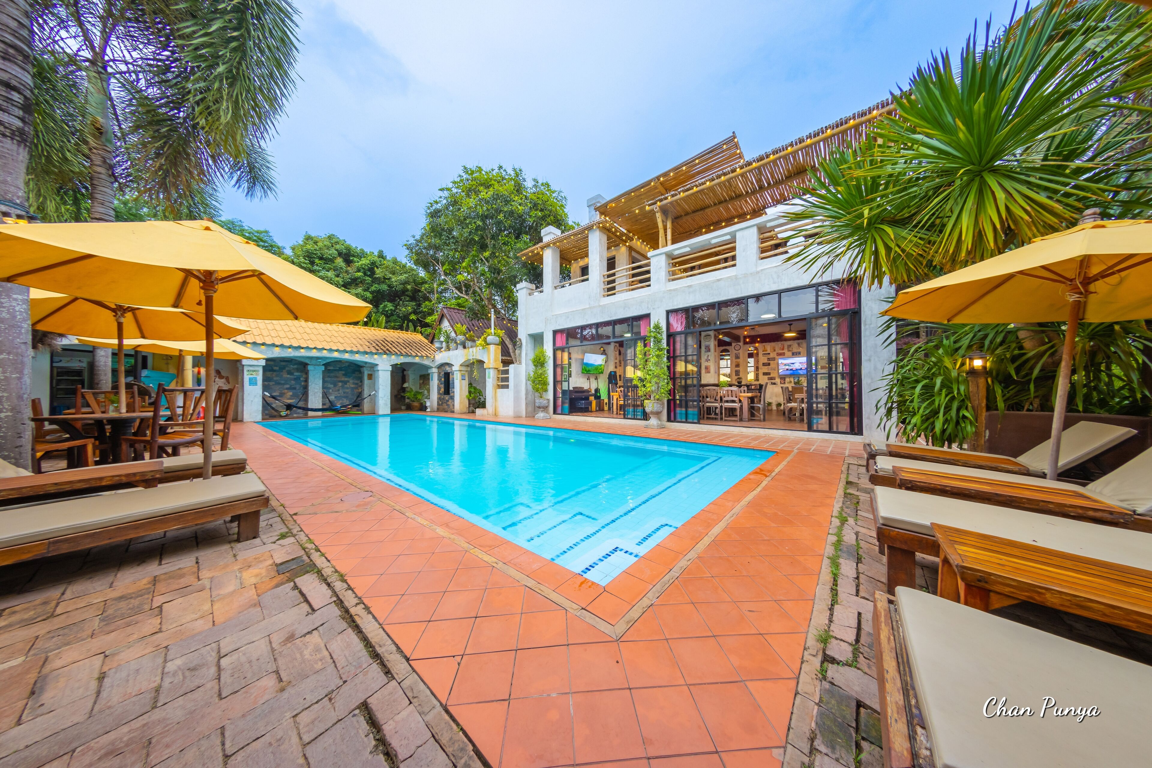 Piscina com espreguiçadeiras de madeira e guarda-sóis amarelos, rodeada por um pátio de tijolos e um edifício com telhado em azulejo.