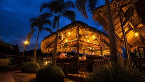 Uma área de bar com telhado de palha, móveis de madeira e luzes de corda, cercada por palmeiras e um caminho de pedra.